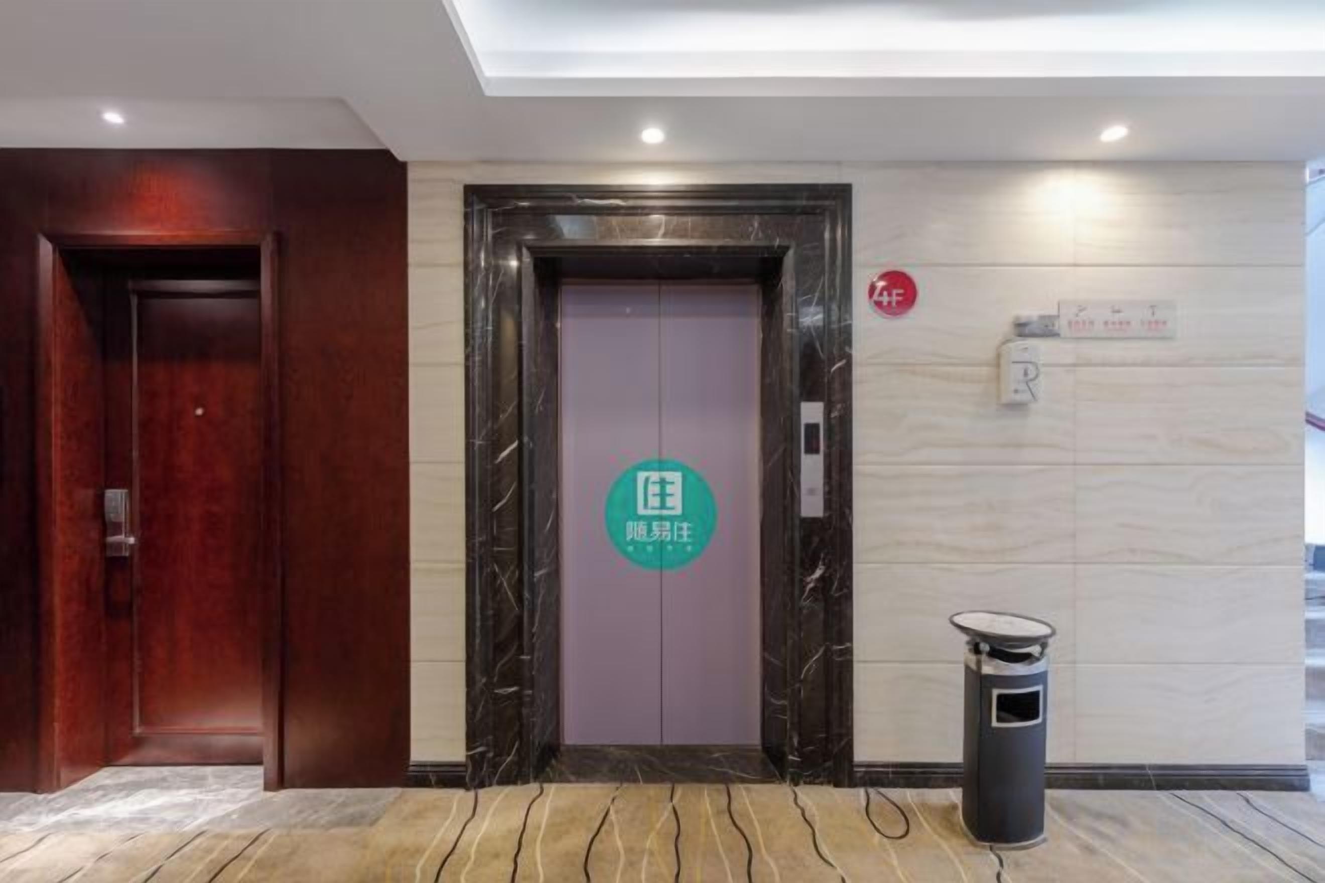 Entrada de hotel com porta de madeira, elevador de mármore, lixeira e placa indicando área de estacionamento.