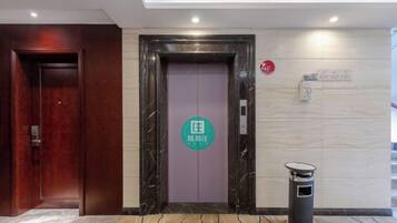Entrada de hotel com porta de madeira, elevador de mármore, lixeira e placa indicando área de estacionamento.