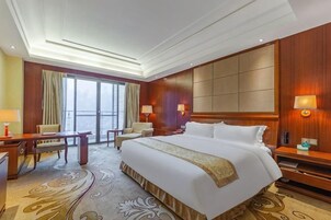 Quarto de hotel com uma cama grande, duas mesas de cabeceira, uma escrivaninha com cadeira, um sofá e uma janela ampla com cortinas.