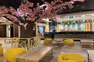 Um lobby com uma grande árvore artificial de cerejeira, bancos de madeira e puffs amarelos.