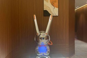 Escultura de vidro transparente com base azul e dois elementos pontudos em laranja, apoiada em uma superfície refletora.