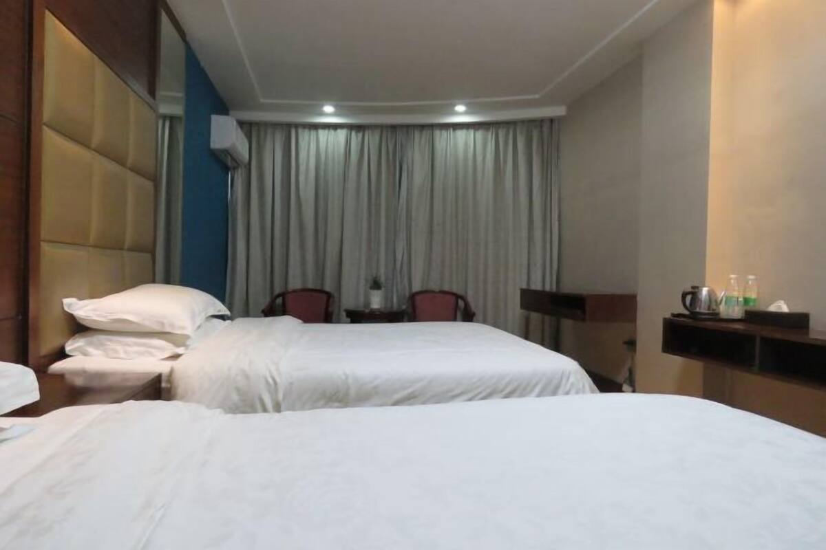 Quarto de hotel com duas camas, uma escrivaninha, cadeiras, uma cortina e uma estante com objetos.
