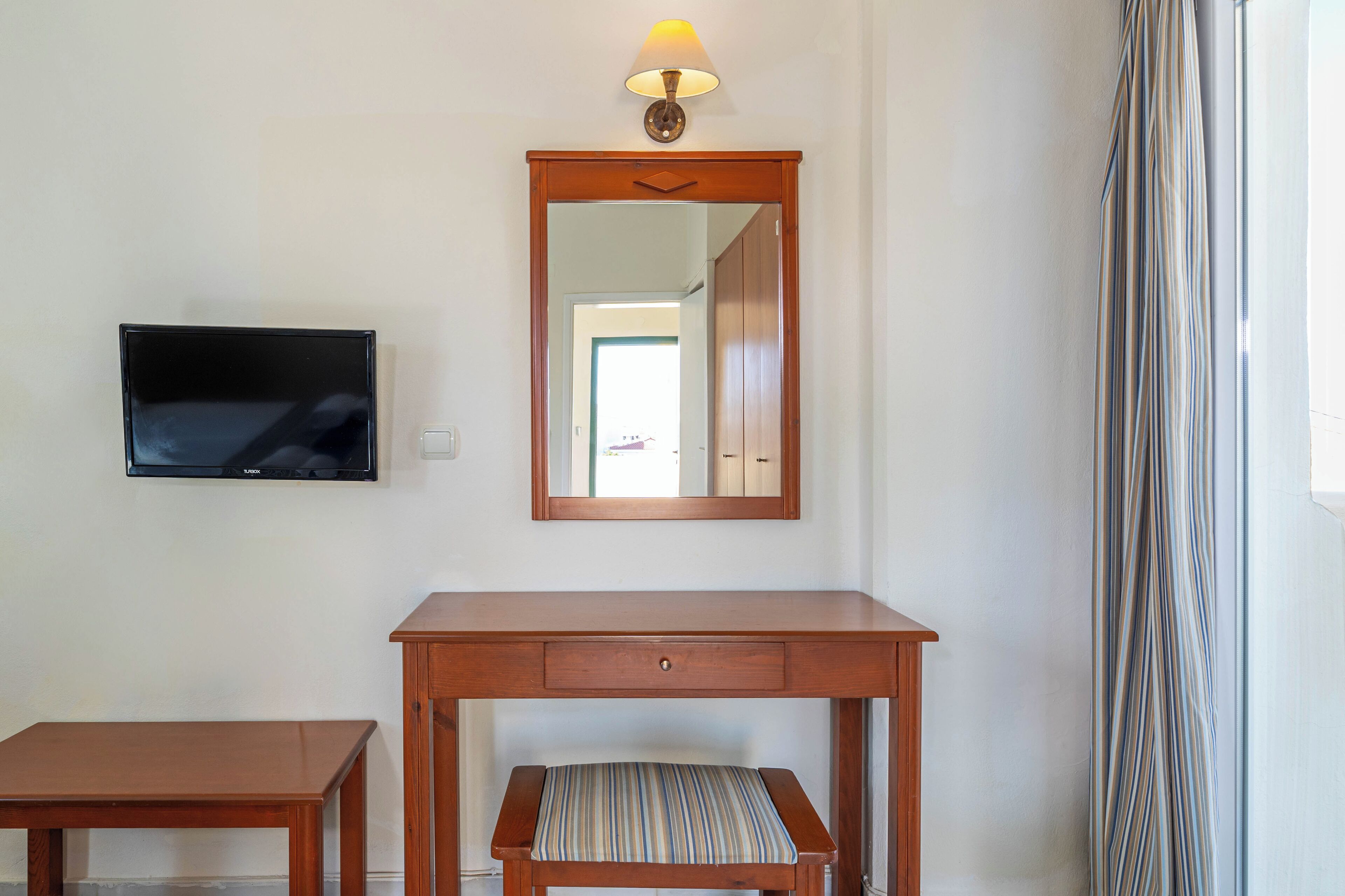 Quarto com escrivaninha de madeira, uma cadeira, televisão na parede e espelho sobre a escrivaninha.