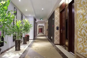 Corredor de hotel com plantas em vasos, piso de madeira e portas de acesso aos quartos.
