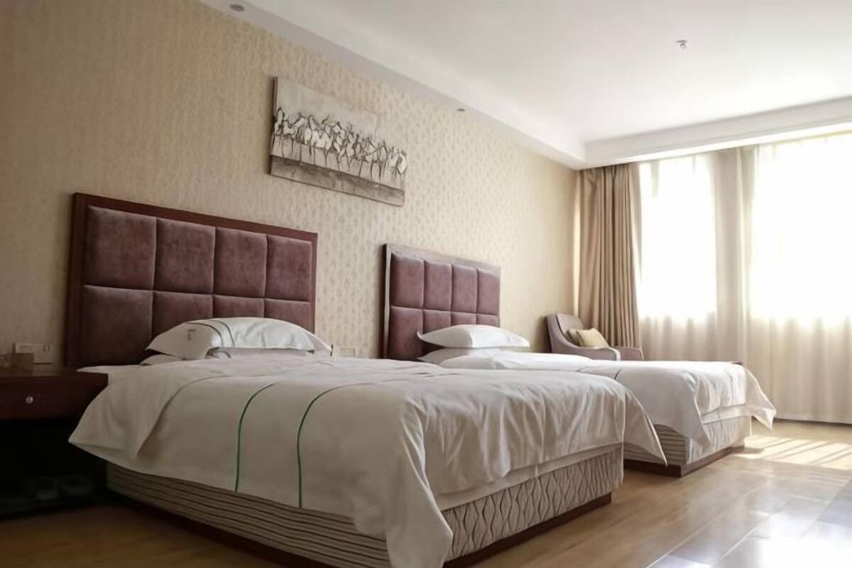 Quarto de hotel com duas camas, uma janela grande com cortinas, uma obra de arte na parede e piso de madeira.