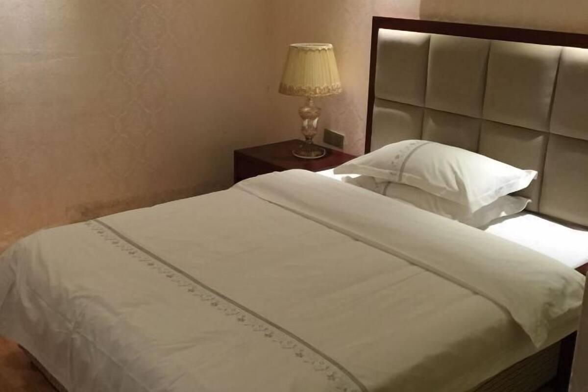 Uma cama bem arrumada com edredom branco e travesseiros, uma luminária de cabeceira e uma mesinha de madeira.