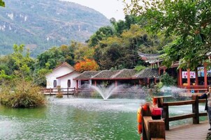 Um pavilhão chinês tradicional sobre a água, com uma passarela de madeira, rodeado por vegetação exuberante e montanhas ao fundo.