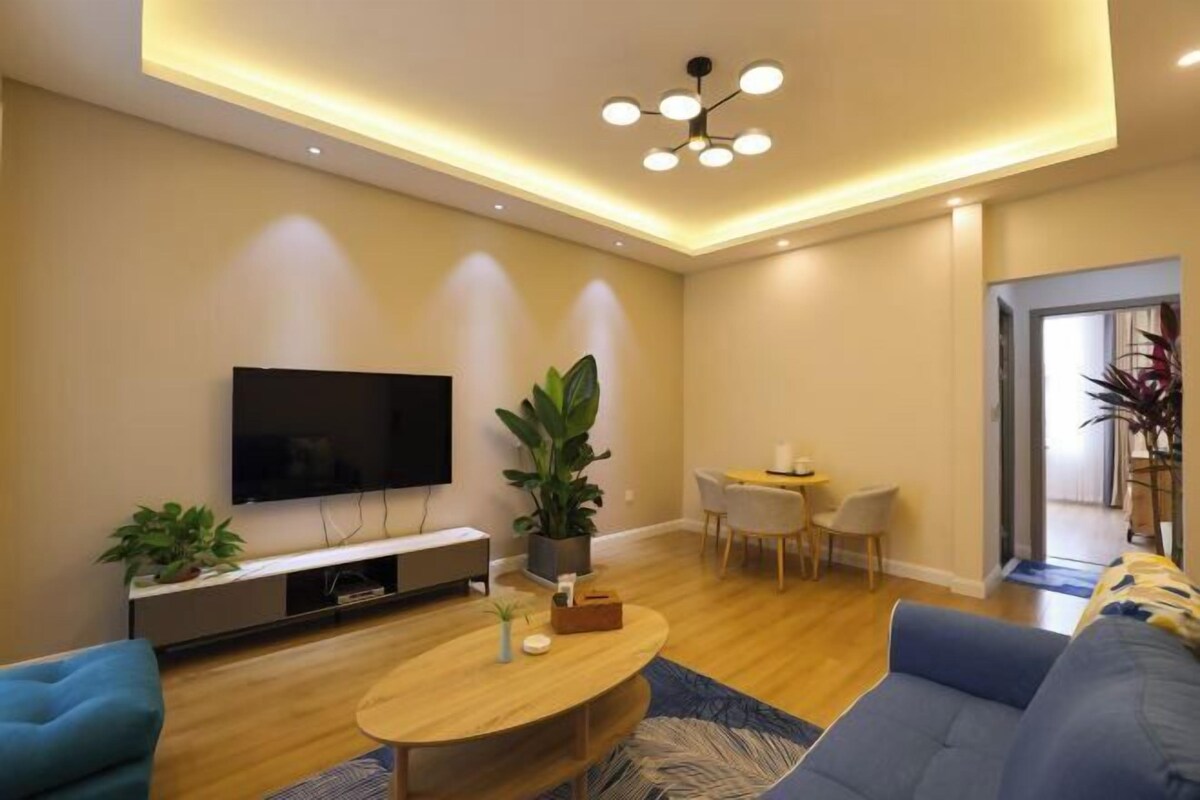 Uma sala de estar moderna com televisão de tela plana, mesa de centro de madeira, sofá azul e um espaço de refeições com uma pequena mesa e cadeiras.