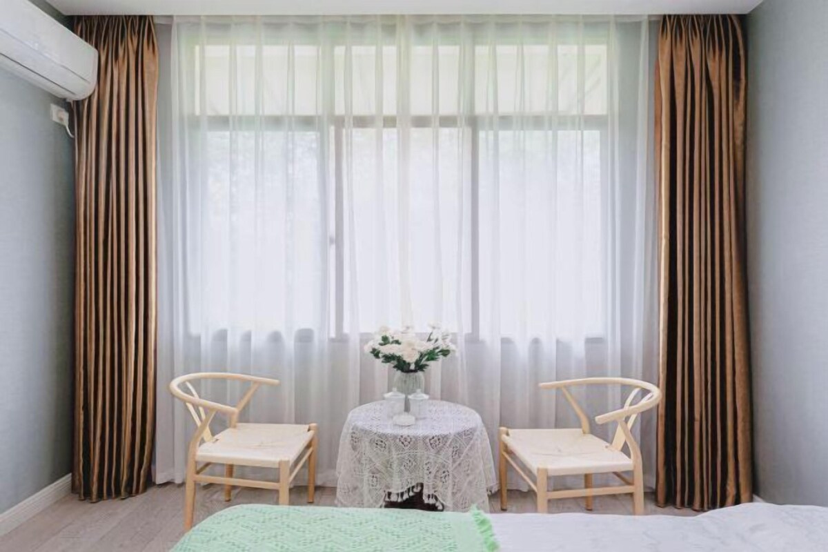 Quarto com cama, duas cadeiras, uma mesinha com um vaso de flores e uma janela com cortinas leves.