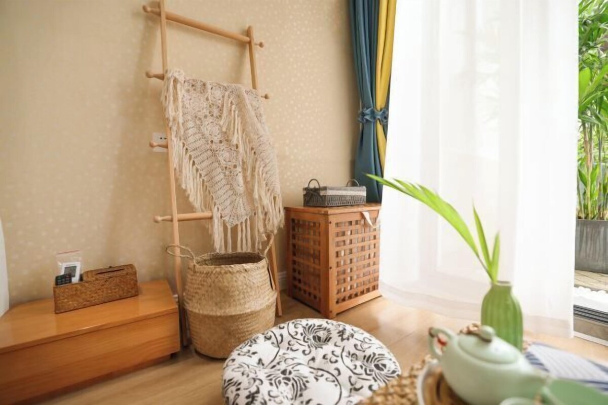 Um quarto aconchegante com piso de madeira, um painel de macramé, uma cesta trançada, um pequeno baú de madeira, um otomano estampado, um vaso verde e uma chaleira.