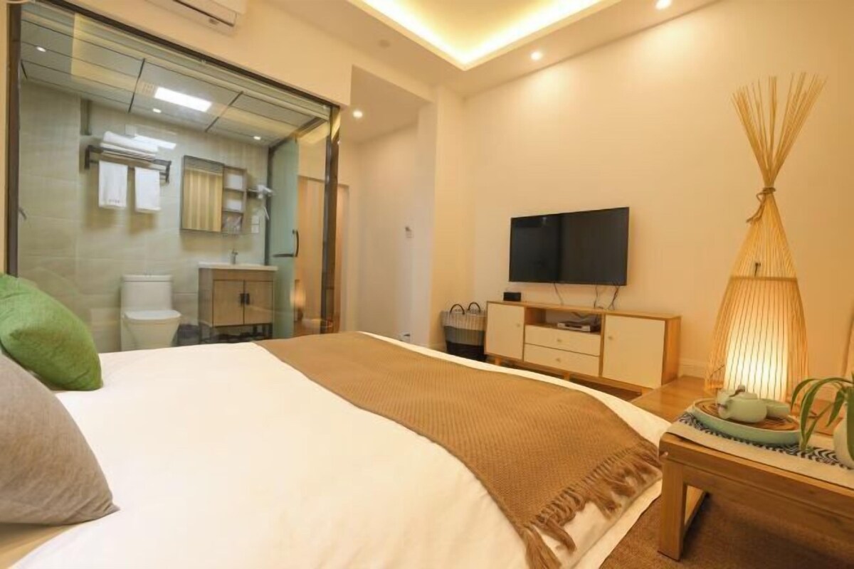 Quarto de hotel moderno com uma cama grande, uma televisão, uma mesinha de cabeceira com abajur e vista para o banheiro através de uma porta de vidro.