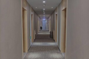 Um corredor longo e estreito com carpete estampado, paredes claras e uma série de portas em um dos lados.