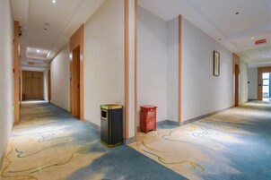 Corredor de hotel com carpete estampado, portas de madeira e uma lixeira.