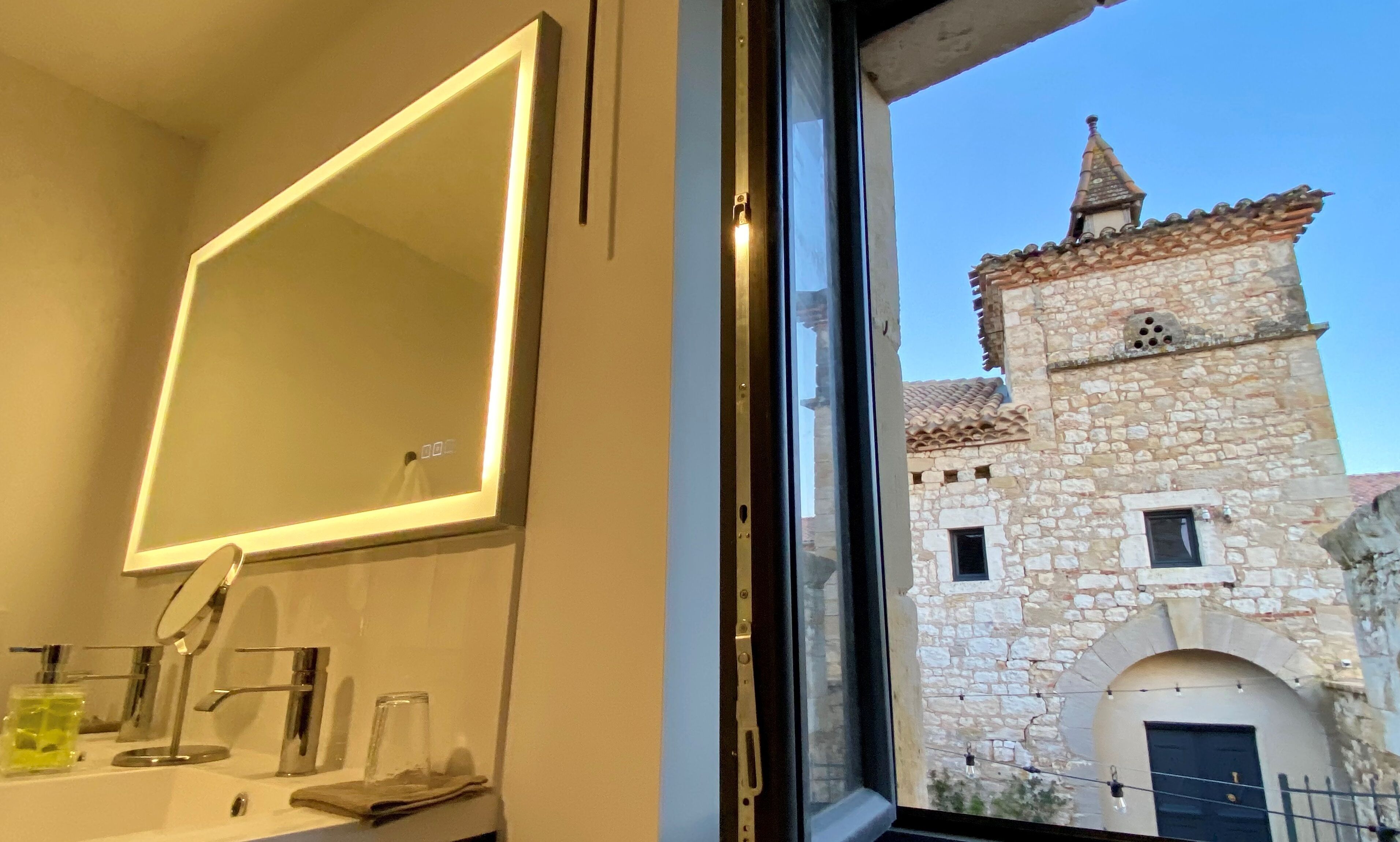 Casa de banho com espelho, lavatório e vista para um edifício de pedra com uma torre.