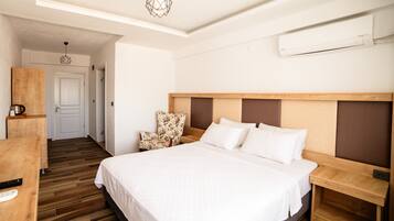 Quarto de hotel com uma cama grande, cabeceira de madeira, uma mesa de cabeceira pequena de madeira, uma cadeira com almofada floral, uma escrivaninha de madeira com uma chaleira e um aparelho de ar condicionado na parede.