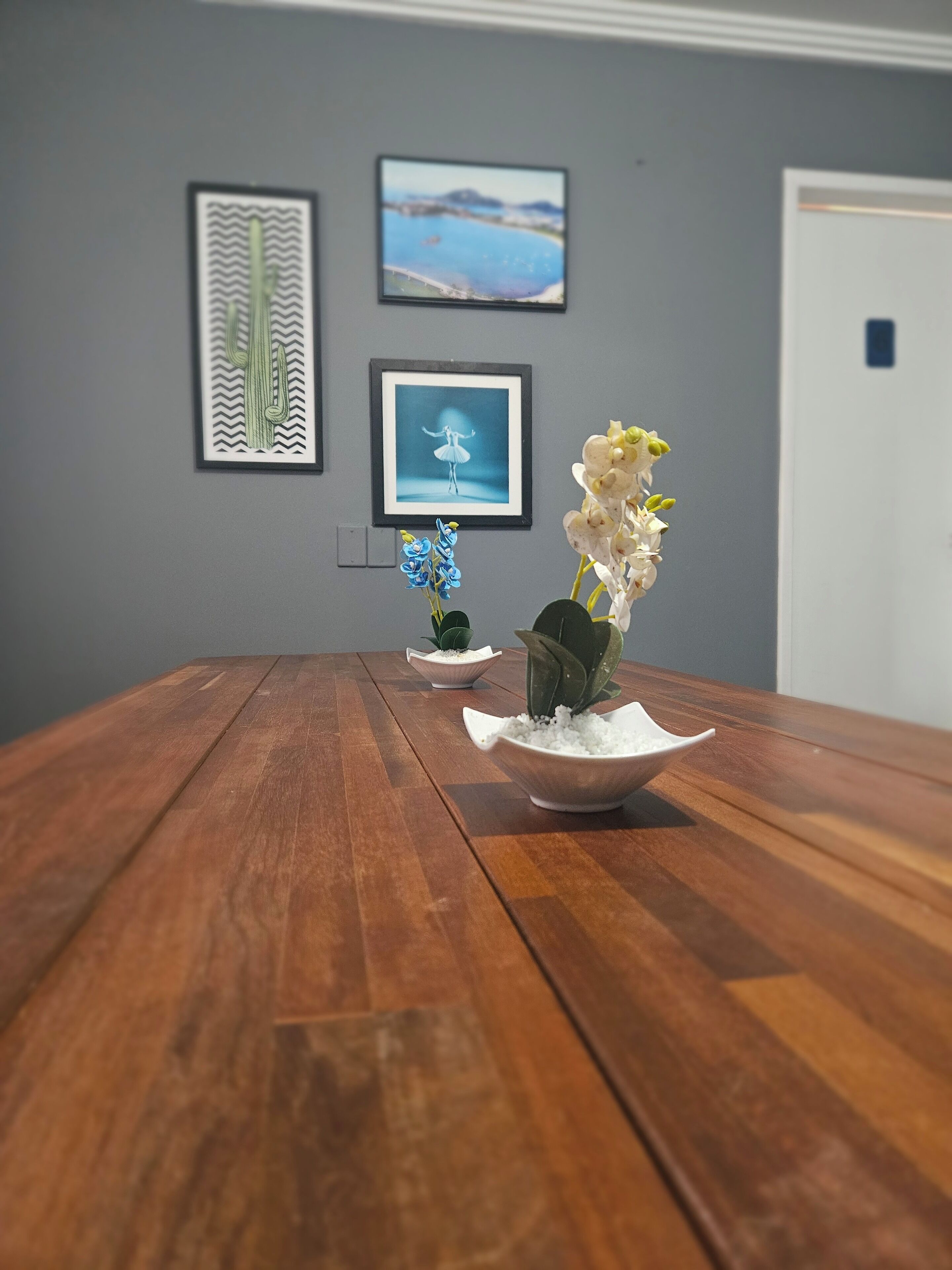 Uma mesa de madeira com três tigelas de cerâmica branca contendo flores brancas, em frente a uma parede cinza com quadros.