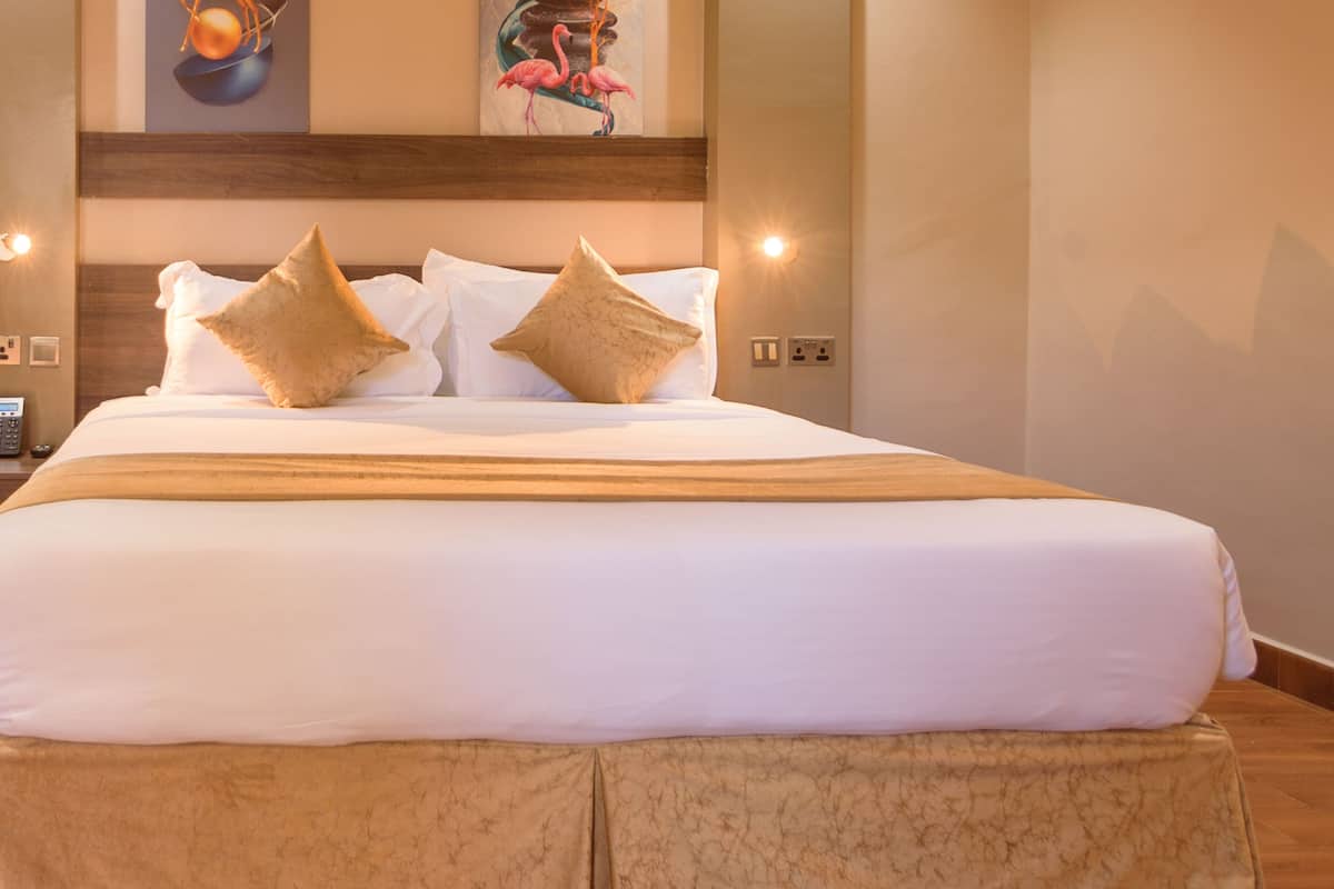 Quarto de hotel com uma cama grande, duas almofadas, cabeceira de madeira, um quadro sobre a cama e uma mesinha de cabeceira com um telefone.