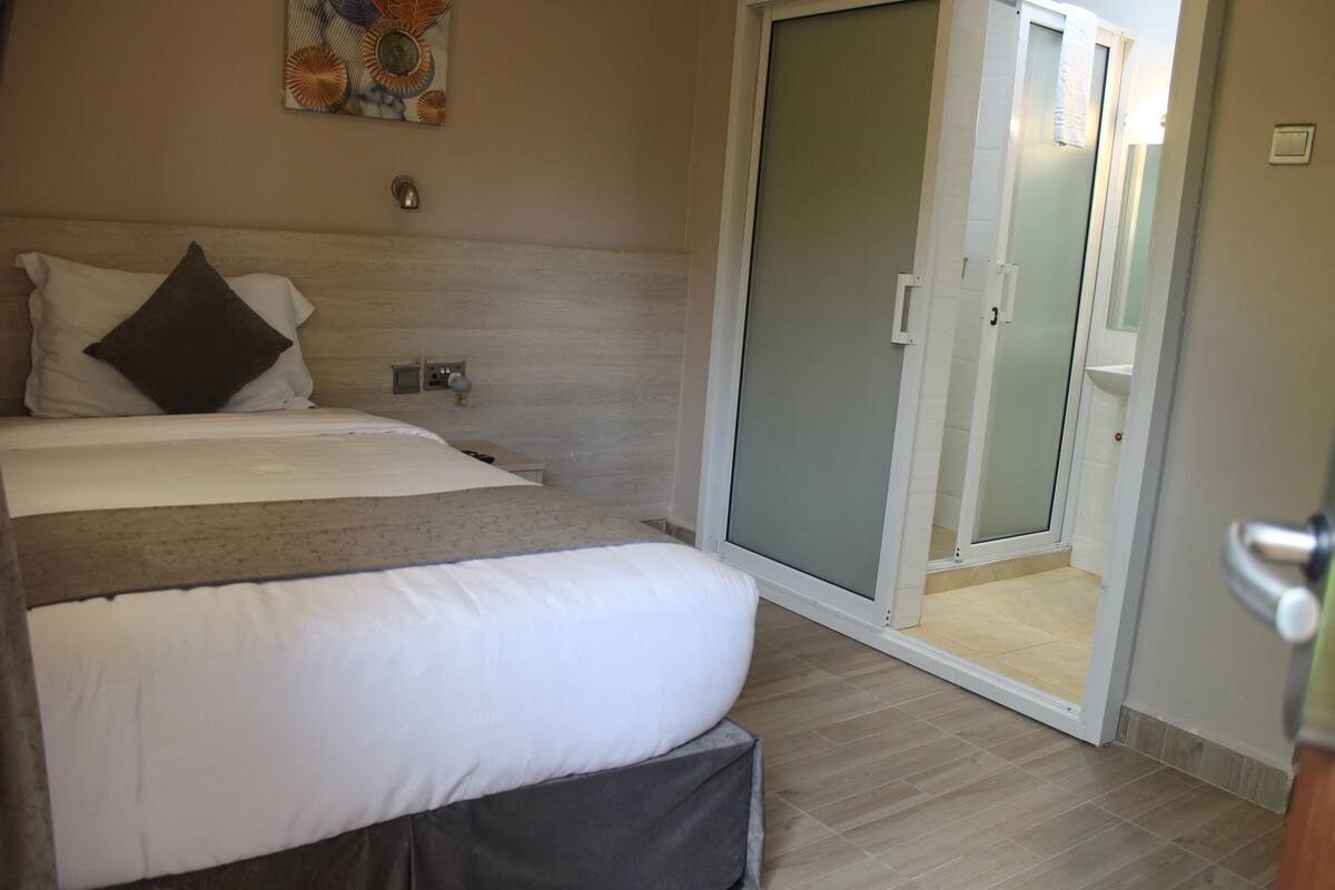 Quarto com cama, mesa de cabeceira, luminária fixada na parede e porta de vidro deslizante que dá para um banheiro.