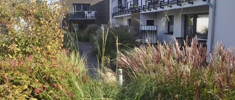 Um edifício moderno de dois andares, com varandas, antena parabólica e um jardim com gramíneas altas e arbustos.