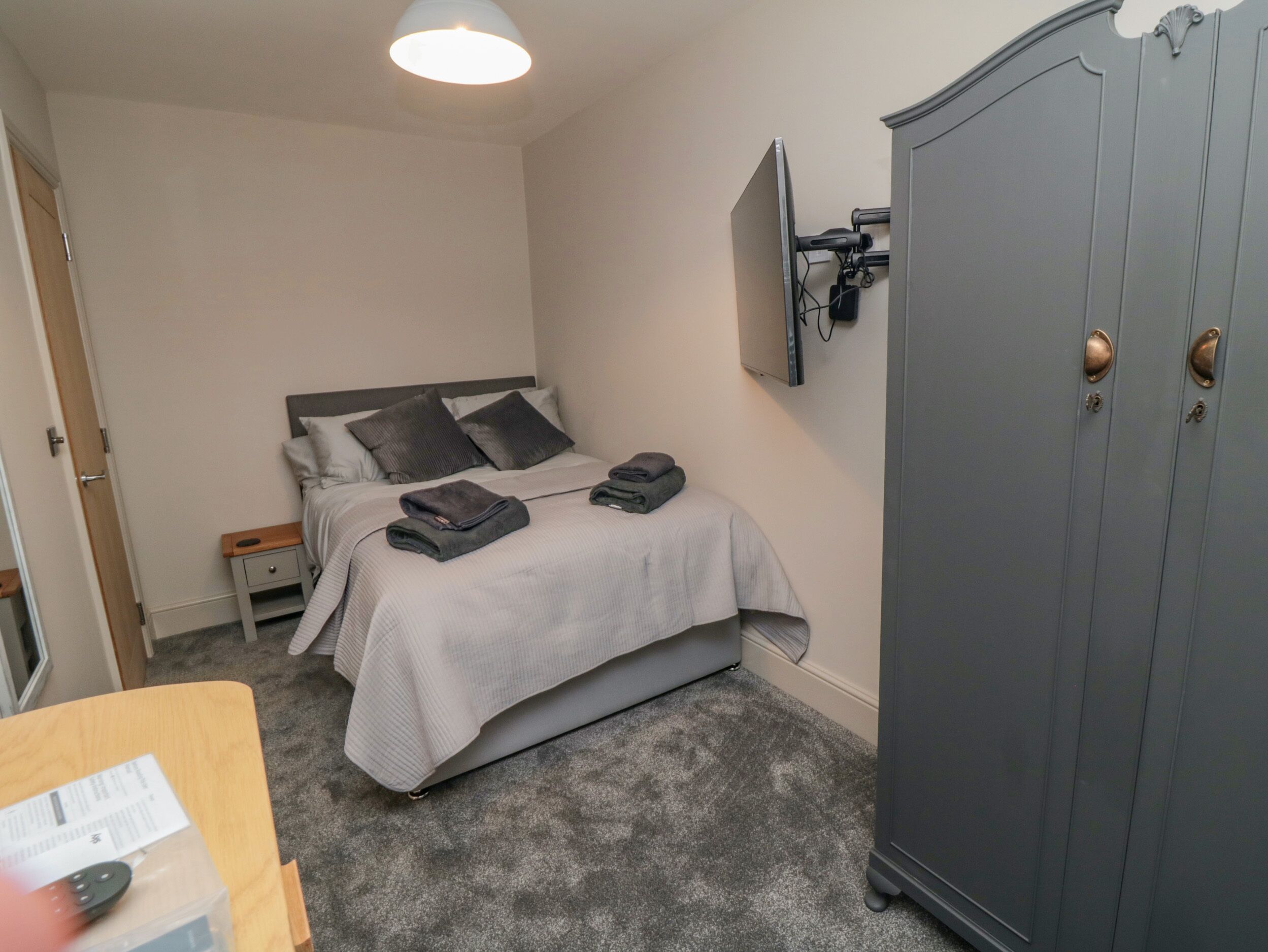 Quarto com cama, guarda-roupa, secretária e televisão fixada na parede.