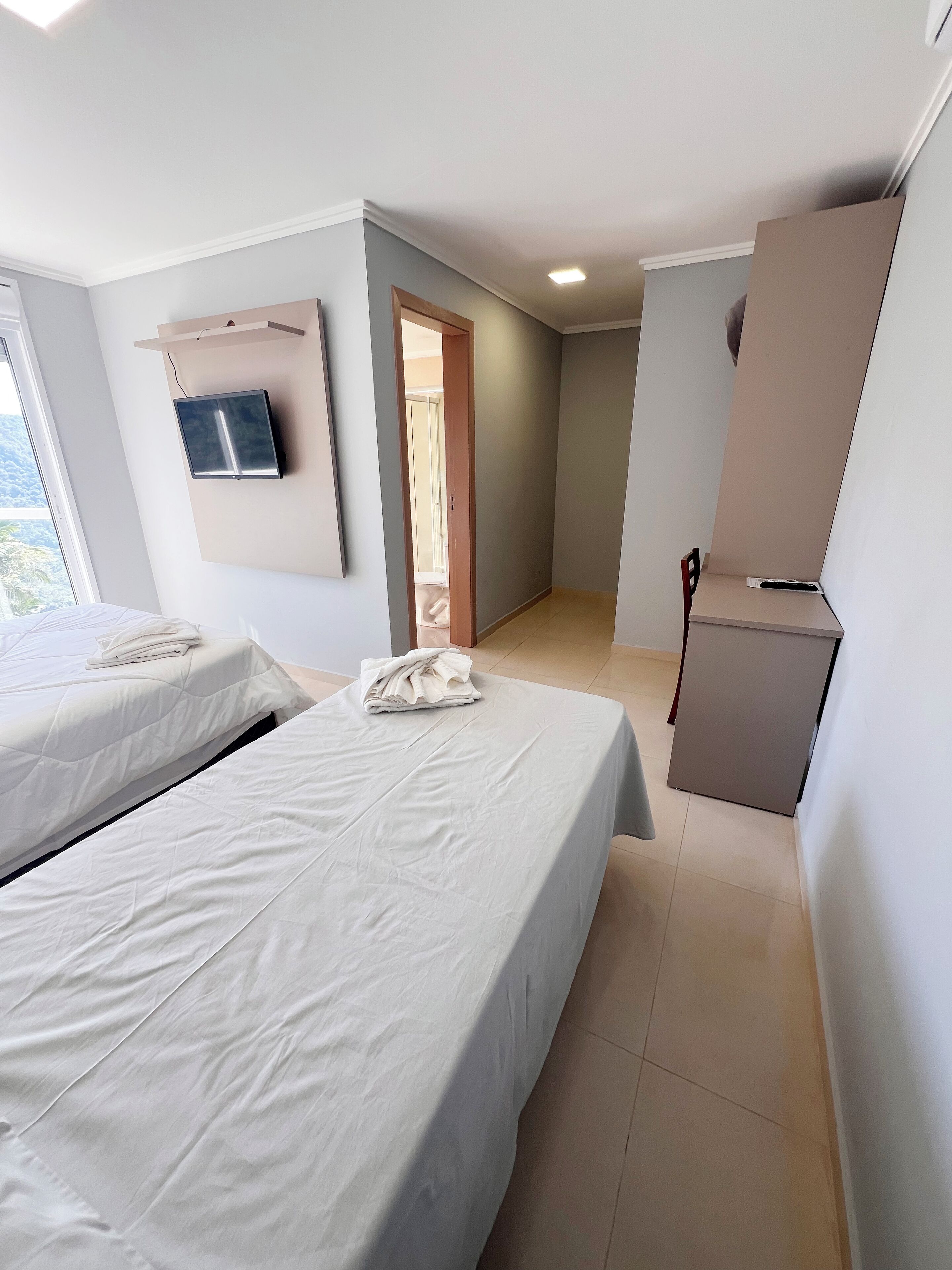 Quarto de hotel com duas camas, uma TV presa à parede e uma pequena escrivaninha com cadeira.