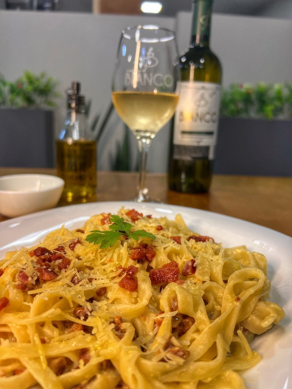 Um prato de massa com bacon e queijo, um copo de vinho branco e uma garrafa de vinho sobre uma mesa.