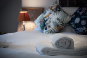 Cama bem arrumada com toalha branca enrolada, abajur vermelho e almofadas com padrão floral.