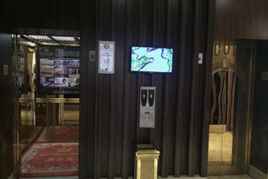 Interior de elevador com televisão na parede, painel de controle e um objeto decorativo no chão.
