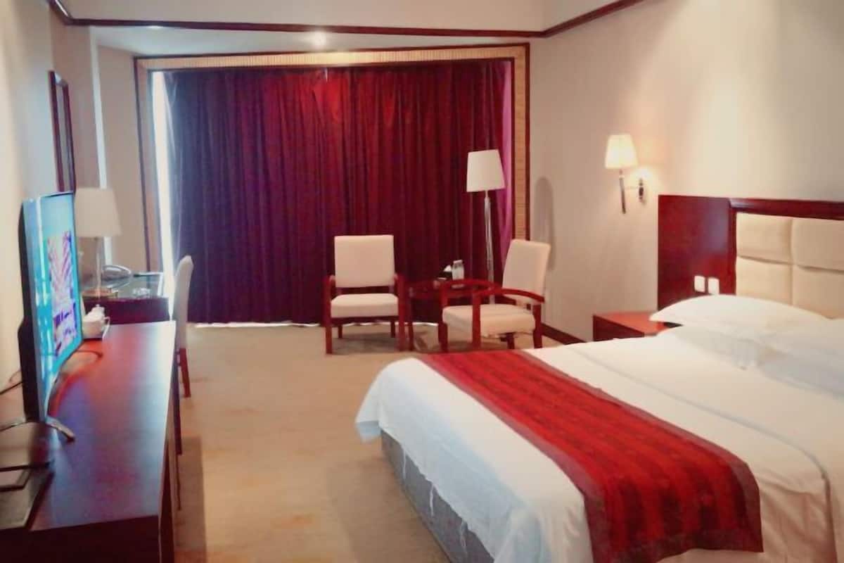 Quarto de hotel com cama, escrivaninha com computador, duas cadeiras, mesa de cabeceira e uma janela grande com cortinas.