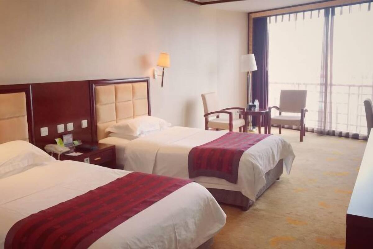 Quarto de hotel com duas camas, uma escrivaninha, uma cadeira e uma janela ampla com vista.