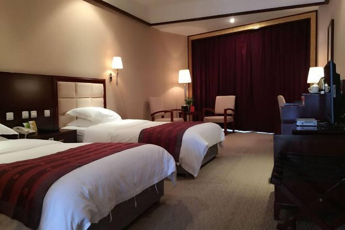 Quarto de hotel com duas camas, uma mesa, uma cadeira, uma televisão e uma janela grande com cortinas.