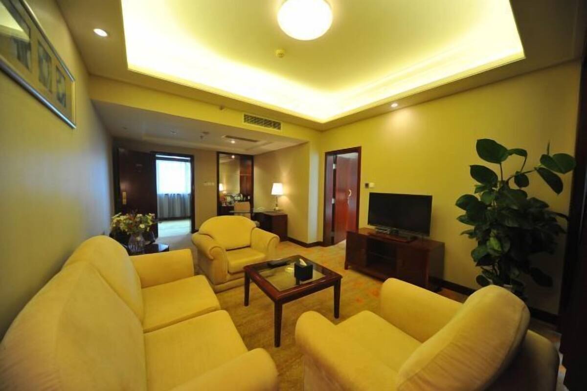 Quarto de hotel com área de estar, incluindo sofás amarelos, uma mesa de centro, uma televisão e uma planta.
