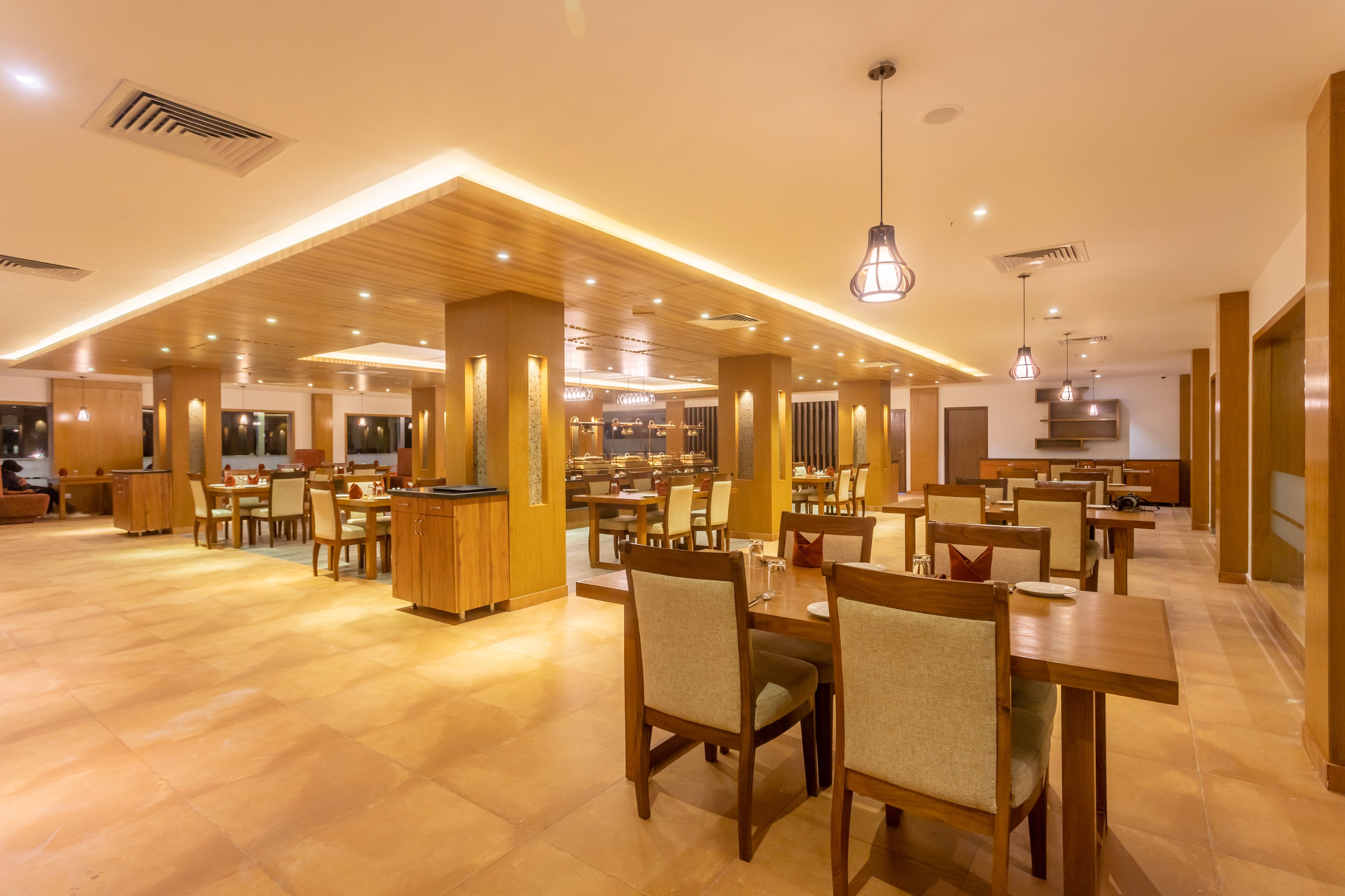 Uma ampla área de refeições de hotel com várias mesas e cadeiras, painéis de madeira nas paredes e iluminação pendente.