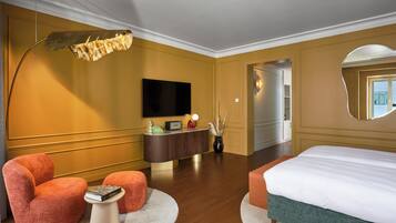 Quarto de hotel com piso de madeira, uma cama grande, uma escrivaninha com televisão de tela plana, uma mesa de centro redonda e duas poltronas laranja.