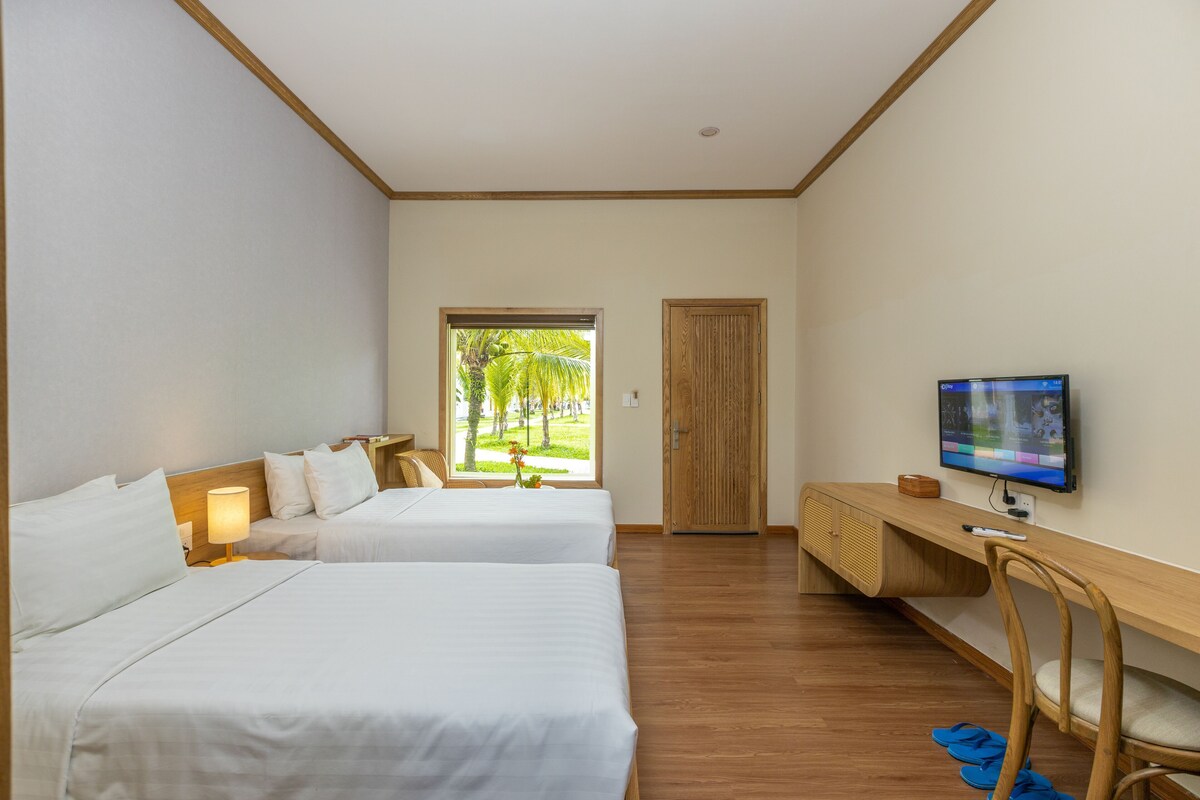 Quarto de hotel com duas camas, uma escrivaninha, uma televisão e vista para um jardim.