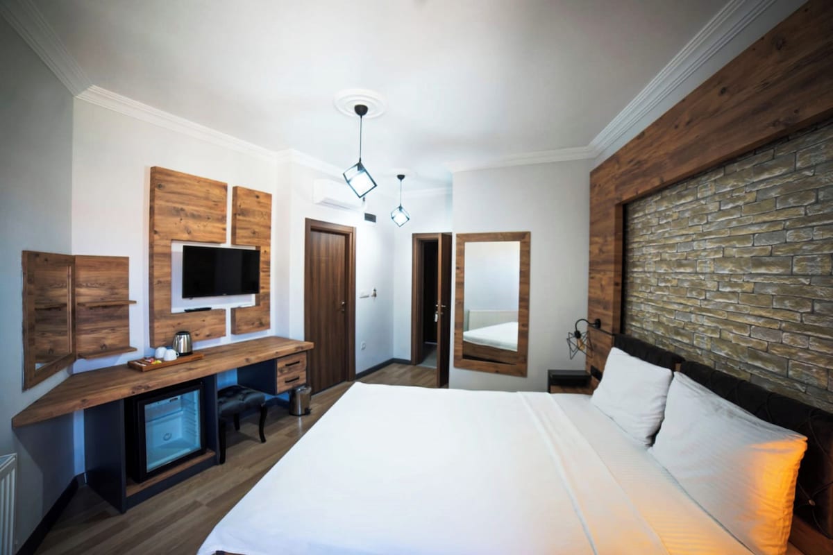 Quarto moderno com cama grande, cabeceira de madeira, televisão integrada, escrivaninha com computador e parede com detalhes em pedra.