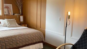 Quarto de hotel com cama, cadeira, escrivaninha, abajur e tapete.