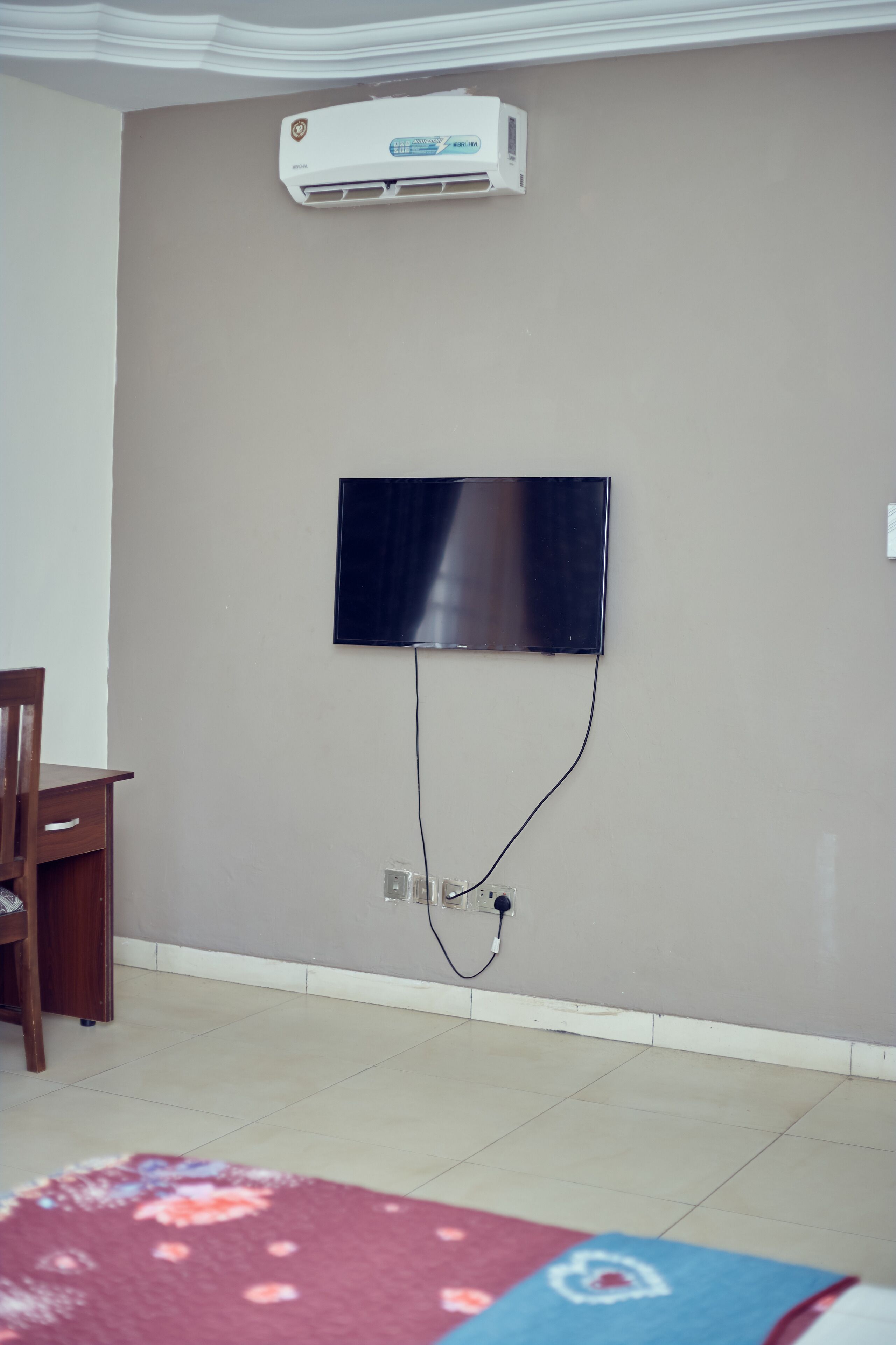 Quarto com televisão de tela plana montada, ar condicionado, cadeira de madeira e cama com colcha estampada.