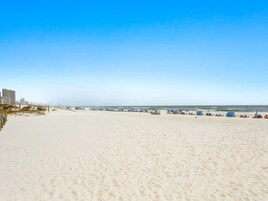 Uma extensa praia de areia com guarda-sóis e espreguiçadeiras, céu claro e edifícios ao fundo.