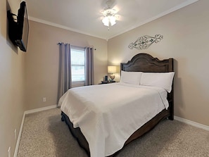 Quarto com cama, ventoinha no teto, televisão fixada na parede, janela com cortinas e um detalhe decorativo na parede.