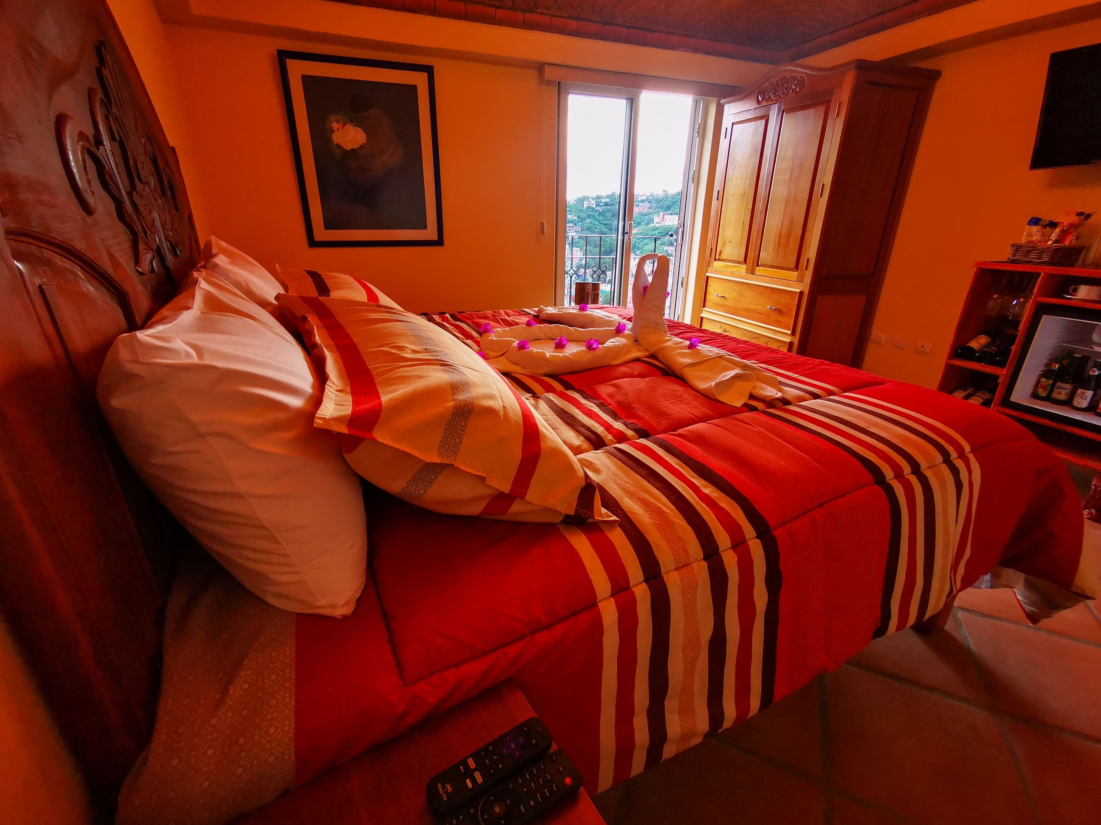 Quarto de hotel com cama, cabeceira de madeira, pintura, janela e minibar.