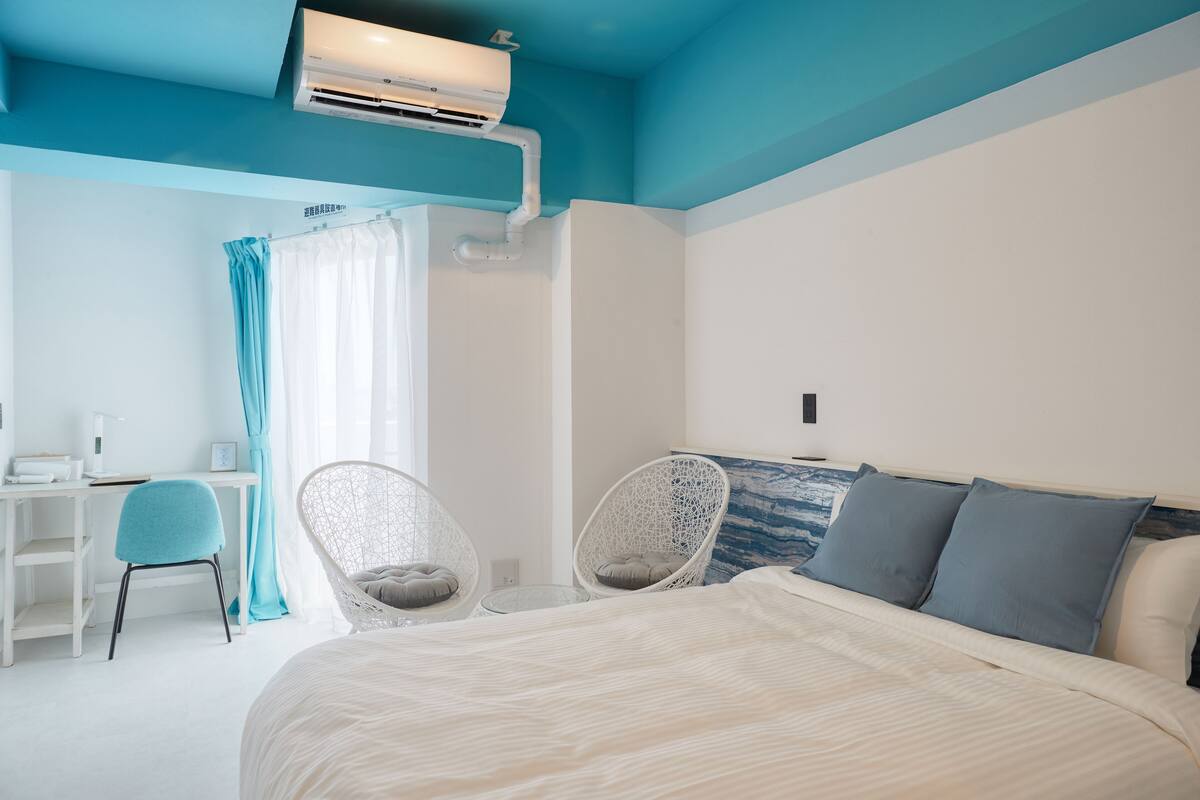 Quarto moderno com uma cama grande, duas cadeiras de vime brancas, uma escrivaninha e um espaço com chuveiro.