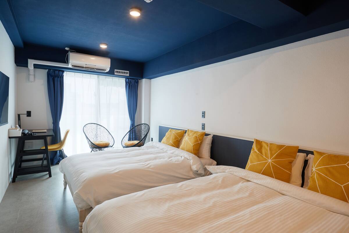 Quarto de hotel com duas camas, uma escrivaninha, uma cadeira e um aparelho de ar condicionado.