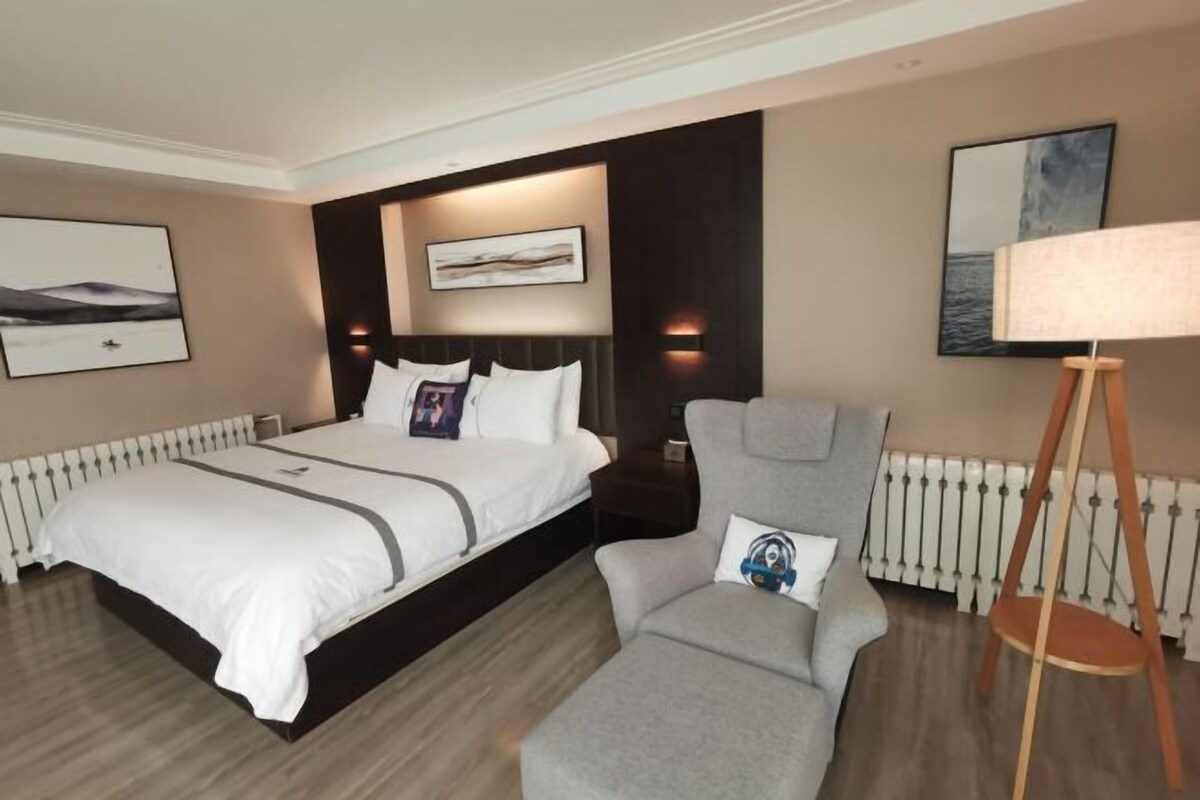 Quarto de hotel com uma cama grande, uma poltrona cinza, um abajur e obras de arte nas paredes.