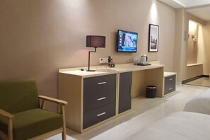Quarto de hotel com cama, uma cadeira verde, escrivaninha com luminária, televisão e um cesto de lixo pequeno.