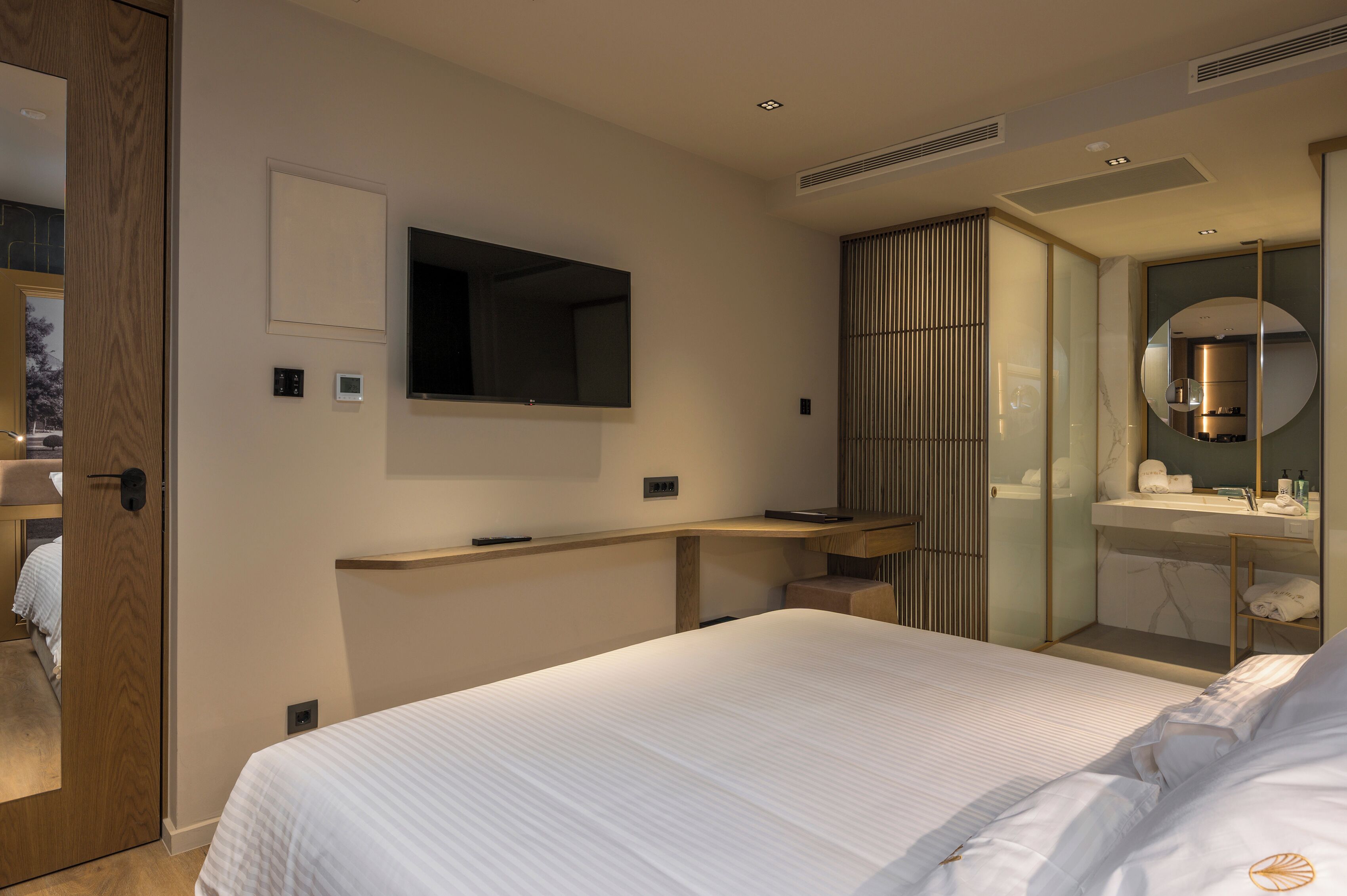Quarto de hotel moderno com uma cama grande, escrivaninha embutida, espelho e banheiro com toucador e pia.