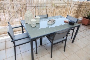 Uma mesa com tampo de vidro, quatro cadeiras, um cercado de bambu trançado e uma planta em vaso ao fundo.