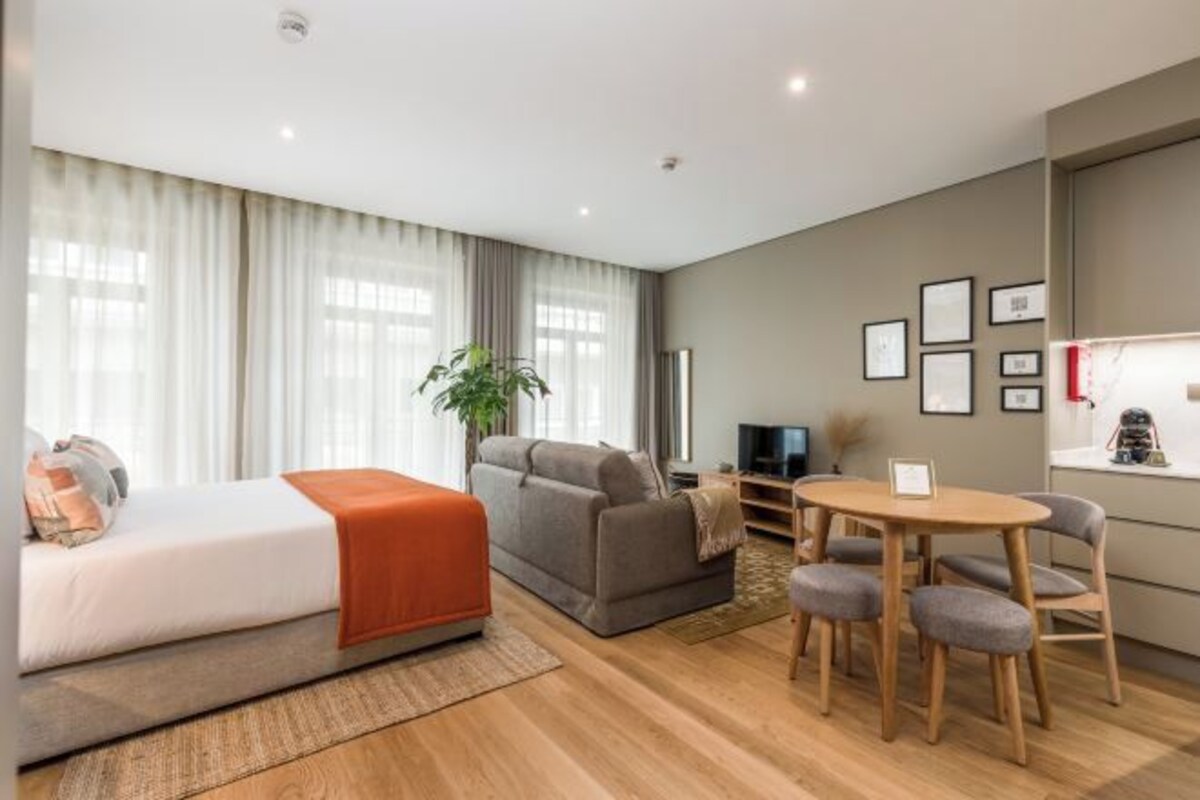 Quarto de hotel moderno com cama, sofá, mesa de jantar com cadeiras, televisão e quadros nas paredes.