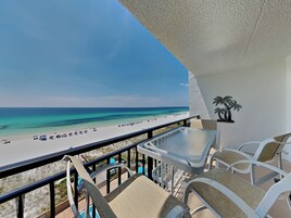 Uma varanda com vista para a praia, uma palmeira decorativa e uma mesa com cadeiras.