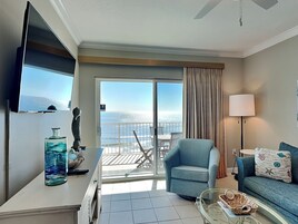Quarto de hotel com varanda, sofá, poltrona, televisão e luminária.